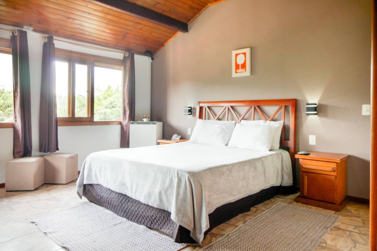 Quarto com cama grande, cabeceira de madeira, mesas de cabeceira e janela com cortinas.