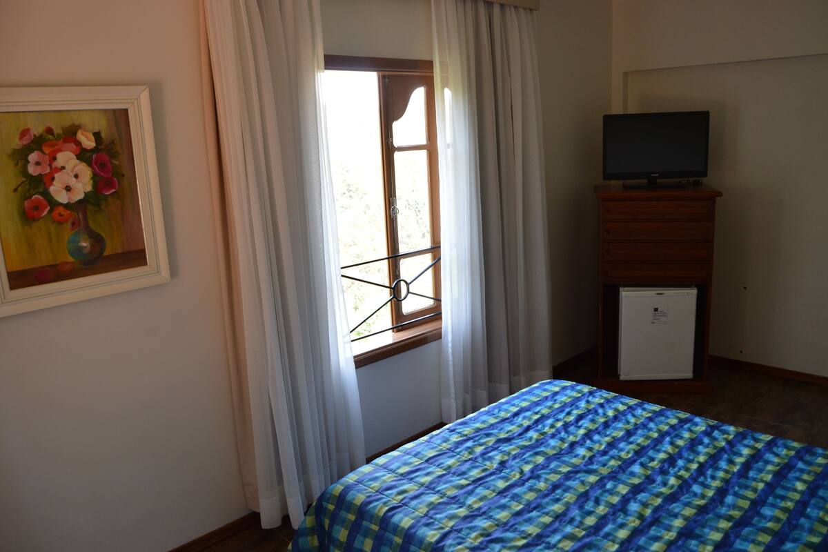 Quarto com cama, televisão em um móvel, um pequeno frigorífico e janela com cortinas.