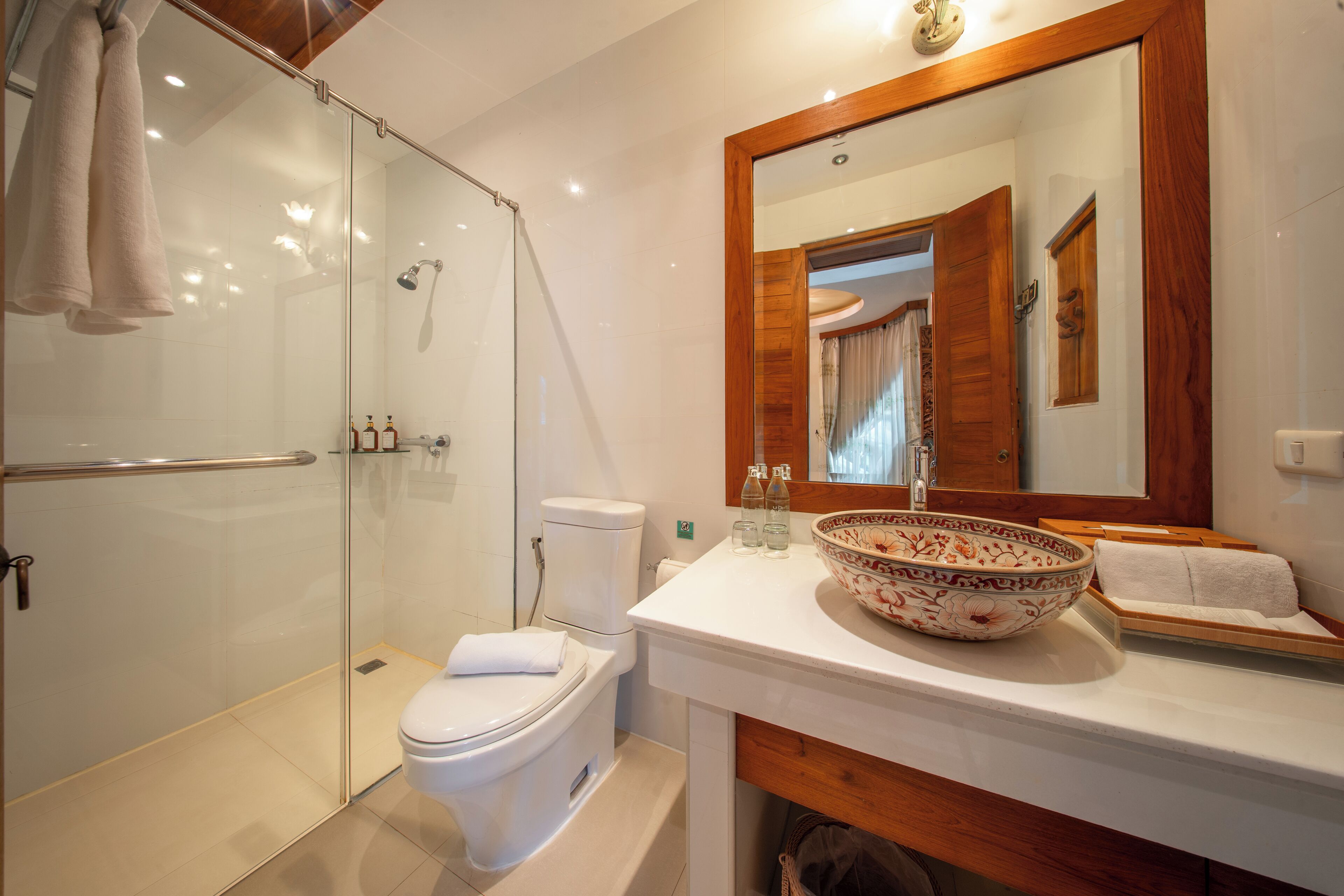 Um banheiro moderno com box de vidro, vaso sanitário branco, toucador de madeira com um espelho grande e uma tigela decorativa no balcão.