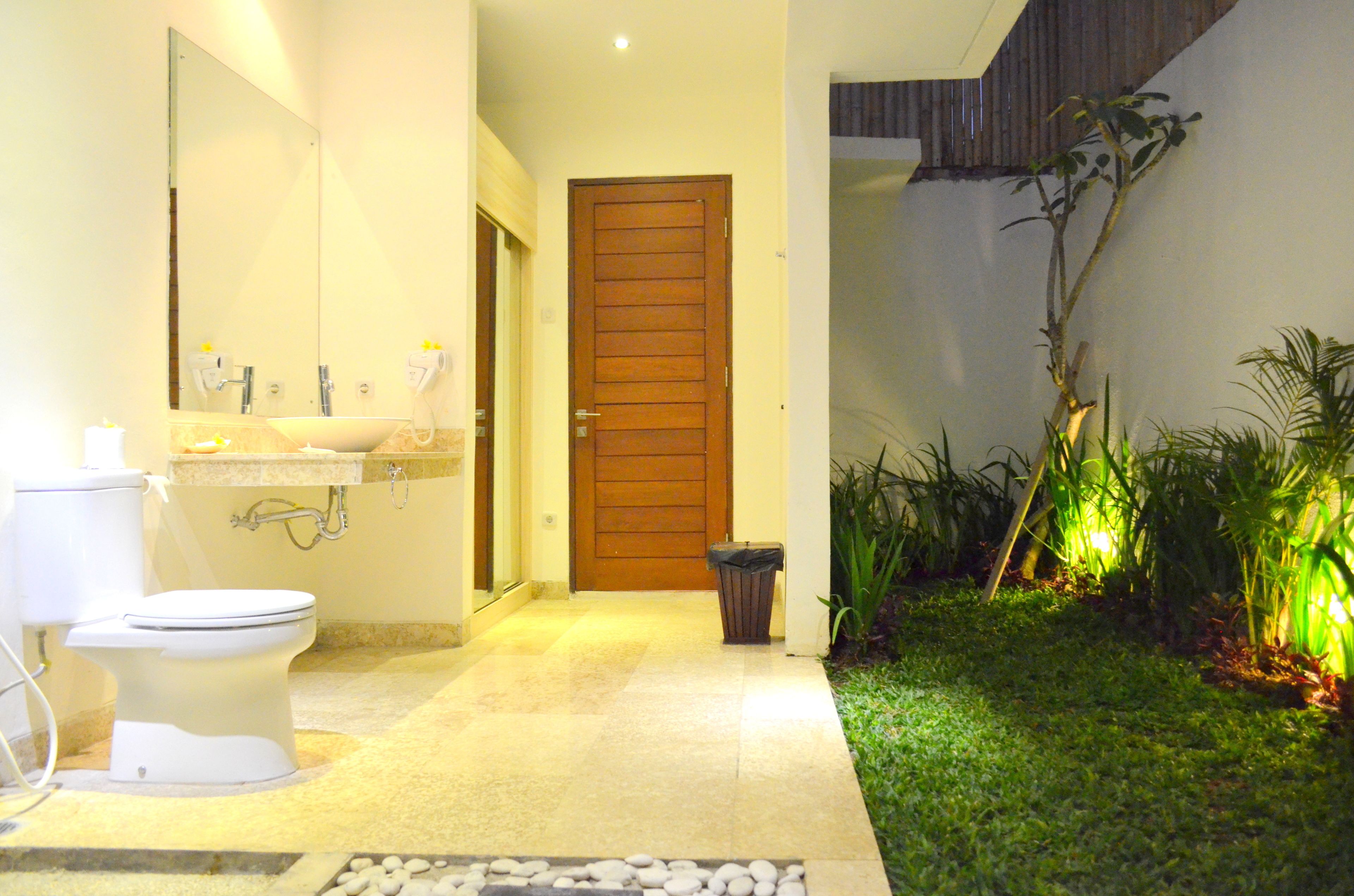 Casa de banho moderna com sanita, lavatório e espelho. Há uma porta que dá para outro quarto e uma pequena área de jardim com plantas e um caminho.