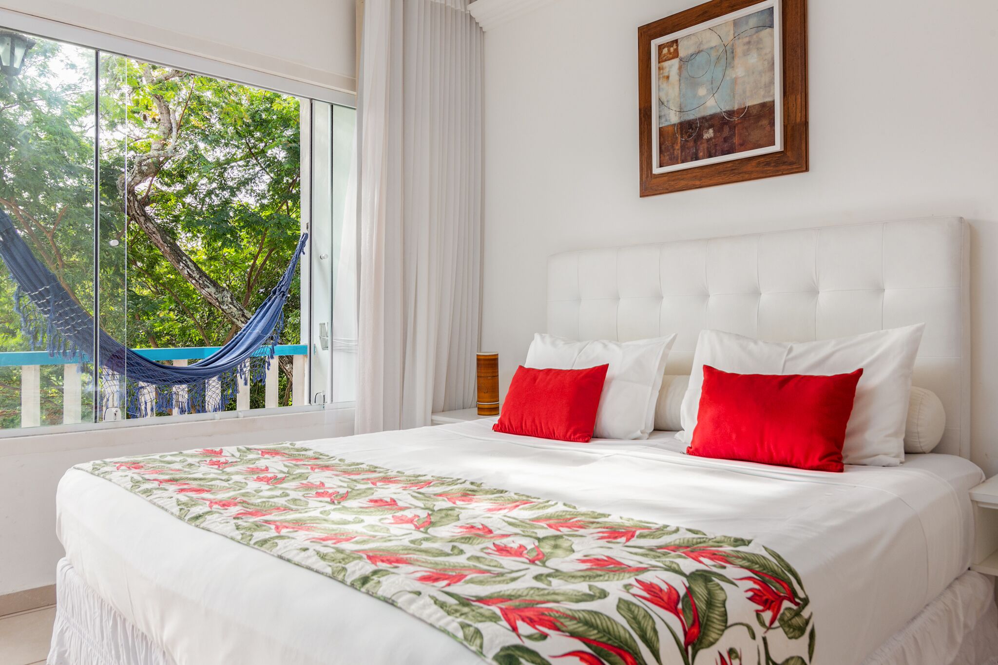 Quarto com cama grande, roupa de cama branca, rede do lado de fora da janela e um quadro na parede.
