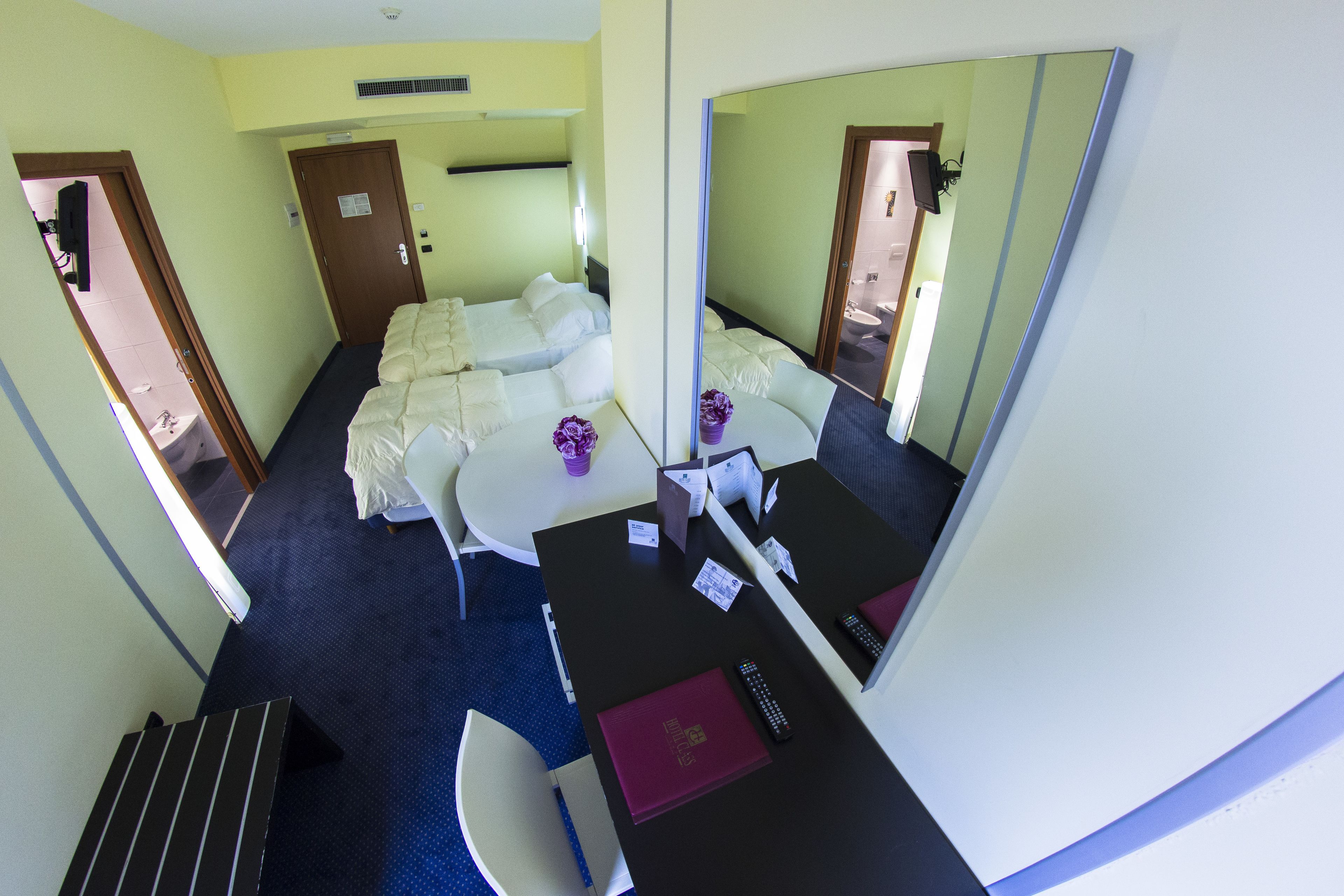 Quarto de hotel com uma cama, uma escrivaninha, uma cadeira, uma mesinha e um espelho.