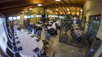 Uma ampla sala de jantar coberta, com várias mesas e cadeiras, vigas de madeira no teto e grandes janelas.