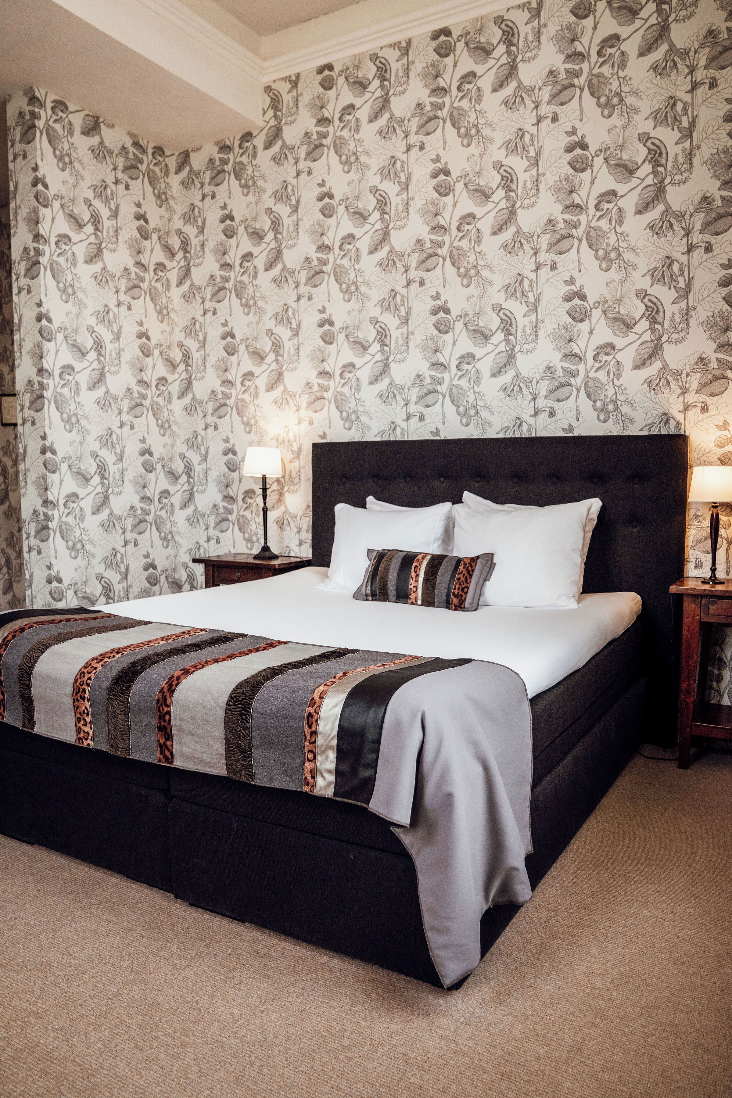 Quarto com cama grande, cabeceira escura, papel de parede floral, duas mesas de cabeceira com abajures e colcha estampada.