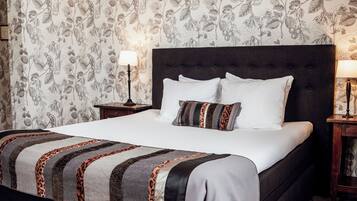 Quarto com cama grande, cabeceira escura, papel de parede floral, duas mesas de cabeceira com abajures e colcha estampada.