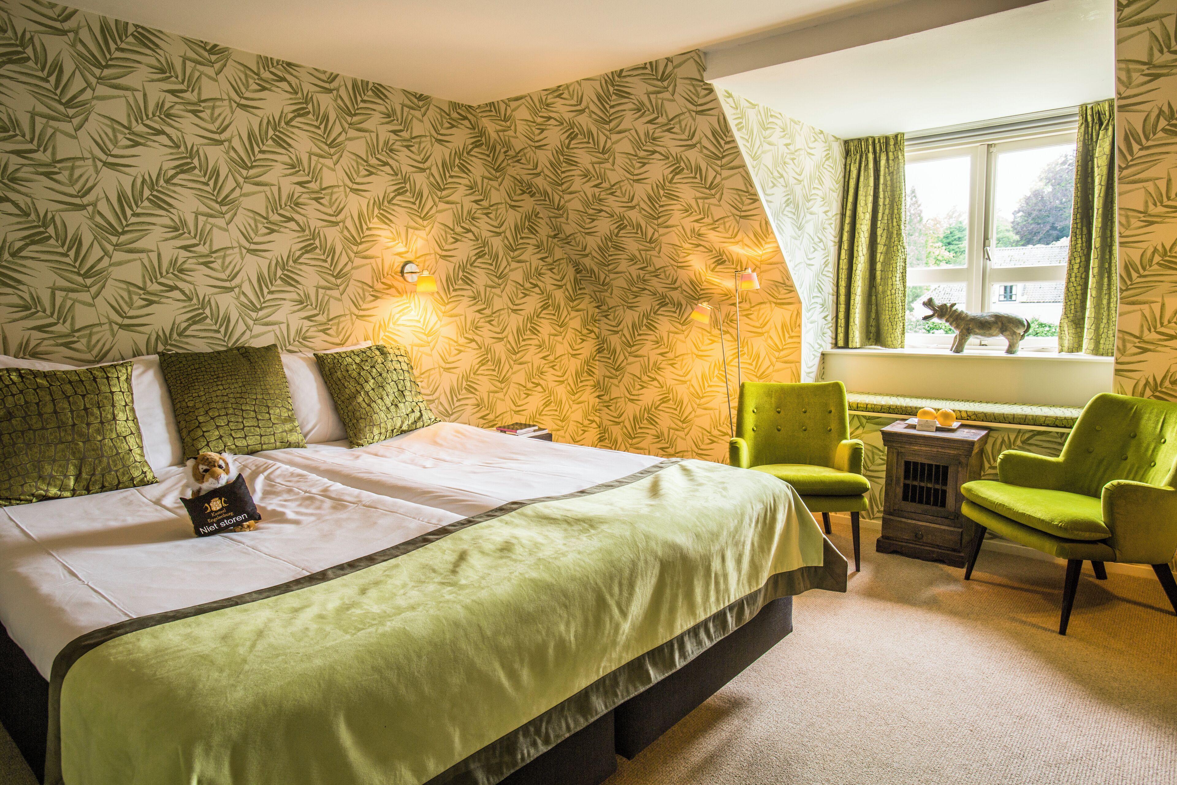 Quarto de hotel com uma cama, cadeiras verdes, uma mesinha e uma janela com vista para um edifício e uma estátua.