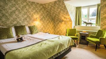 Quarto de hotel com uma cama, cadeiras verdes, uma mesinha e uma janela com vista para um edifício e uma estátua.