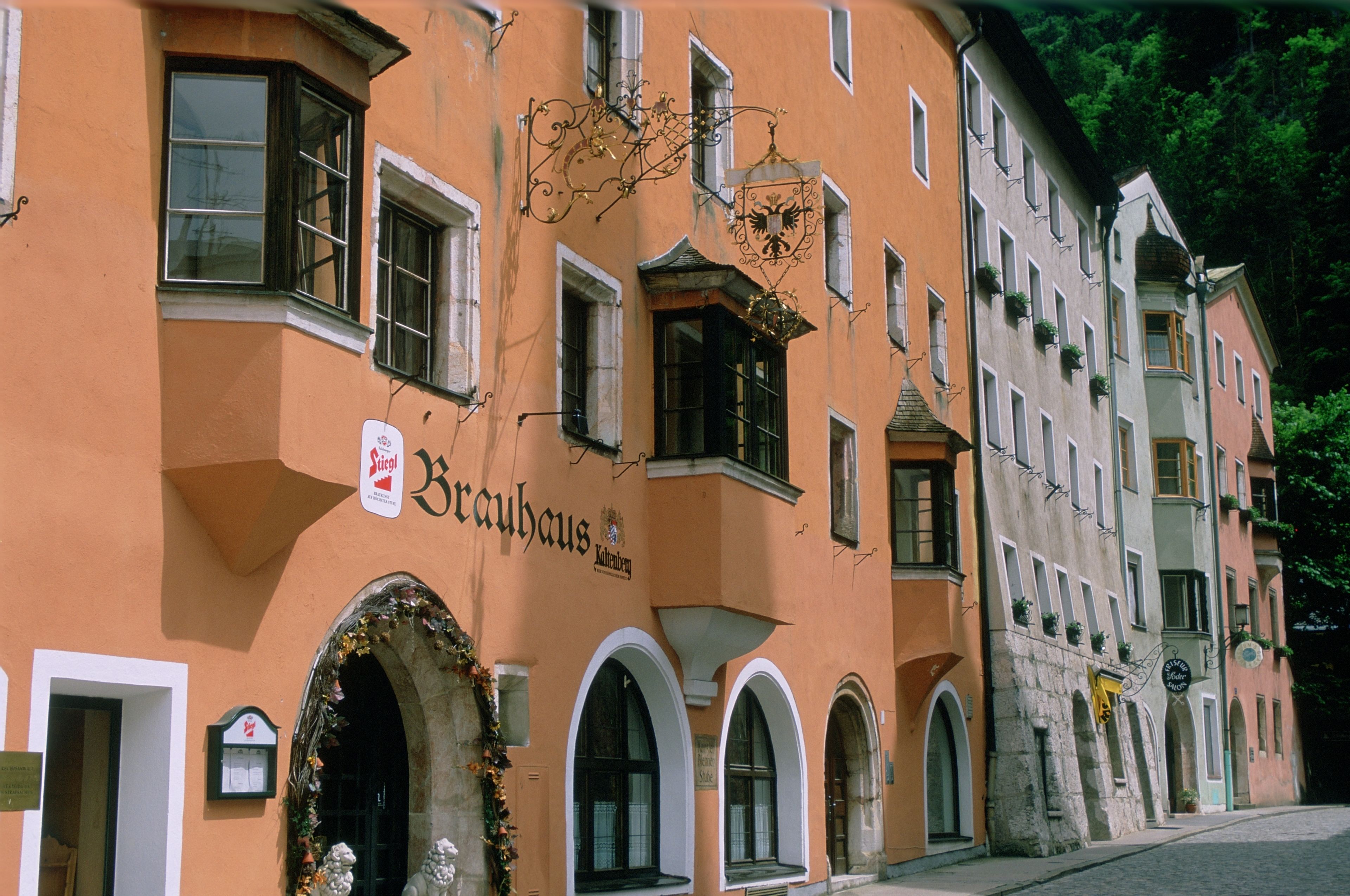 Um edifício com a placa 'Brauhaus', indicando que se trata de uma cervejaria. Possui várias janelas, algumas com toldos, e uma entrada em arco de pedra.