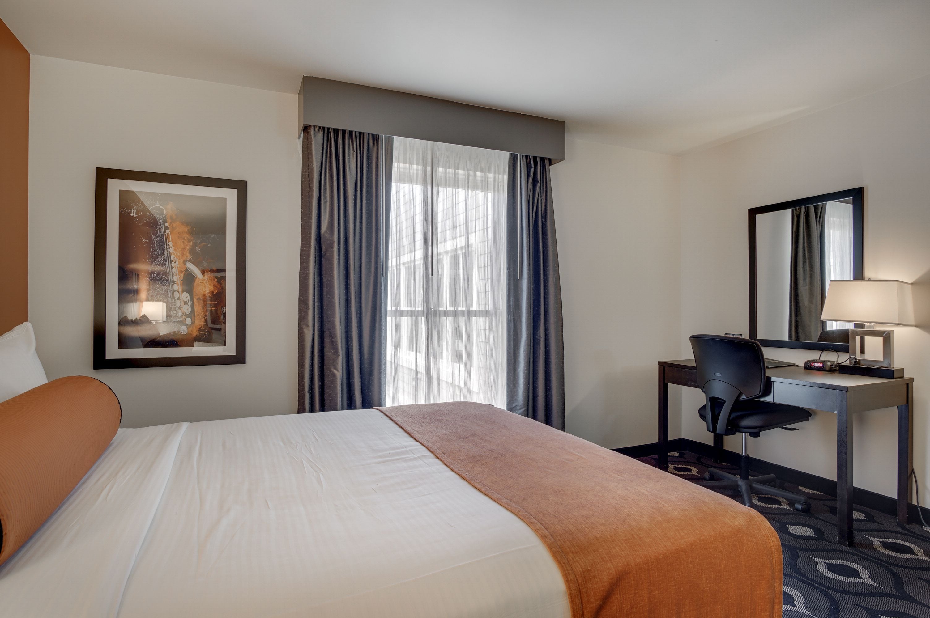 Quarto de hotel com cama, escrivaninha, cadeira, abajur, espelho e janela com cortinas.