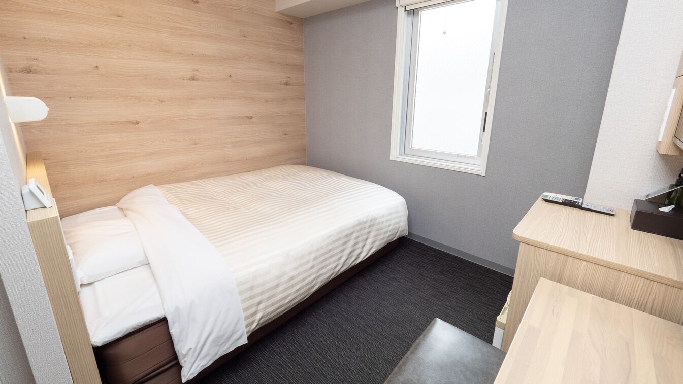 Quarto de hotel compacto com uma cama de solteiro, uma escrivaninha pequena, uma janela e uma parede cinzenta.