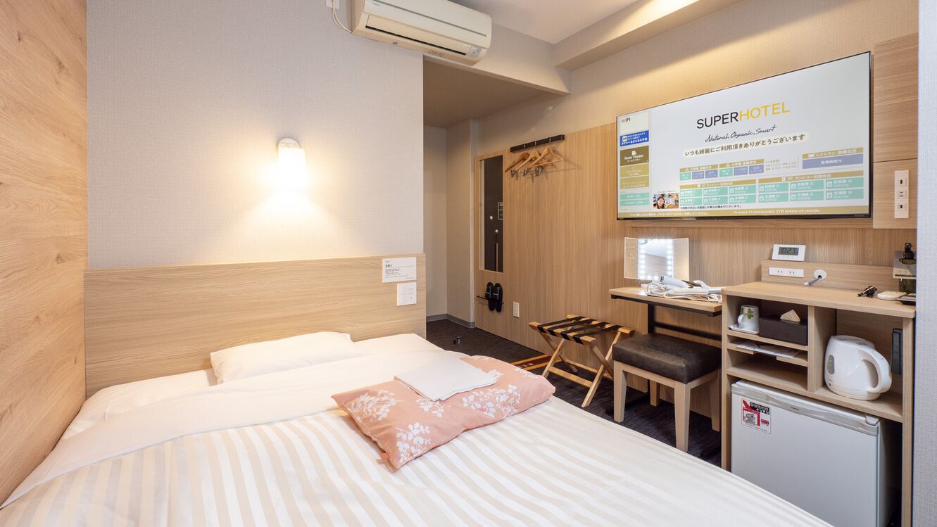 Quarto de hotel com duas camas, uma escrivaninha, uma cadeira, um mini refrigerador e uma televisão na parede exibindo informações do hotel.