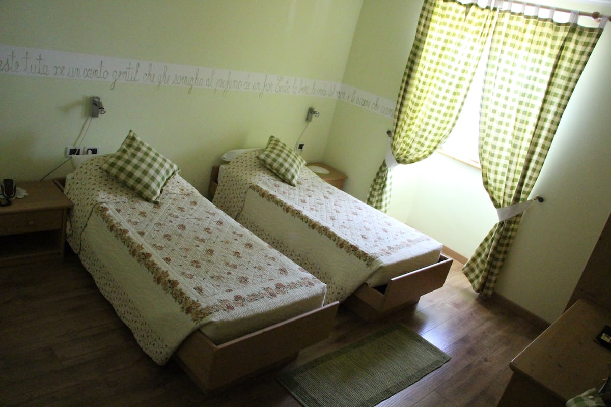 Quarto com duas camas de solteiro, uma mesinha de cabeceira com abajur, tomada na parede e janela com cortinas.