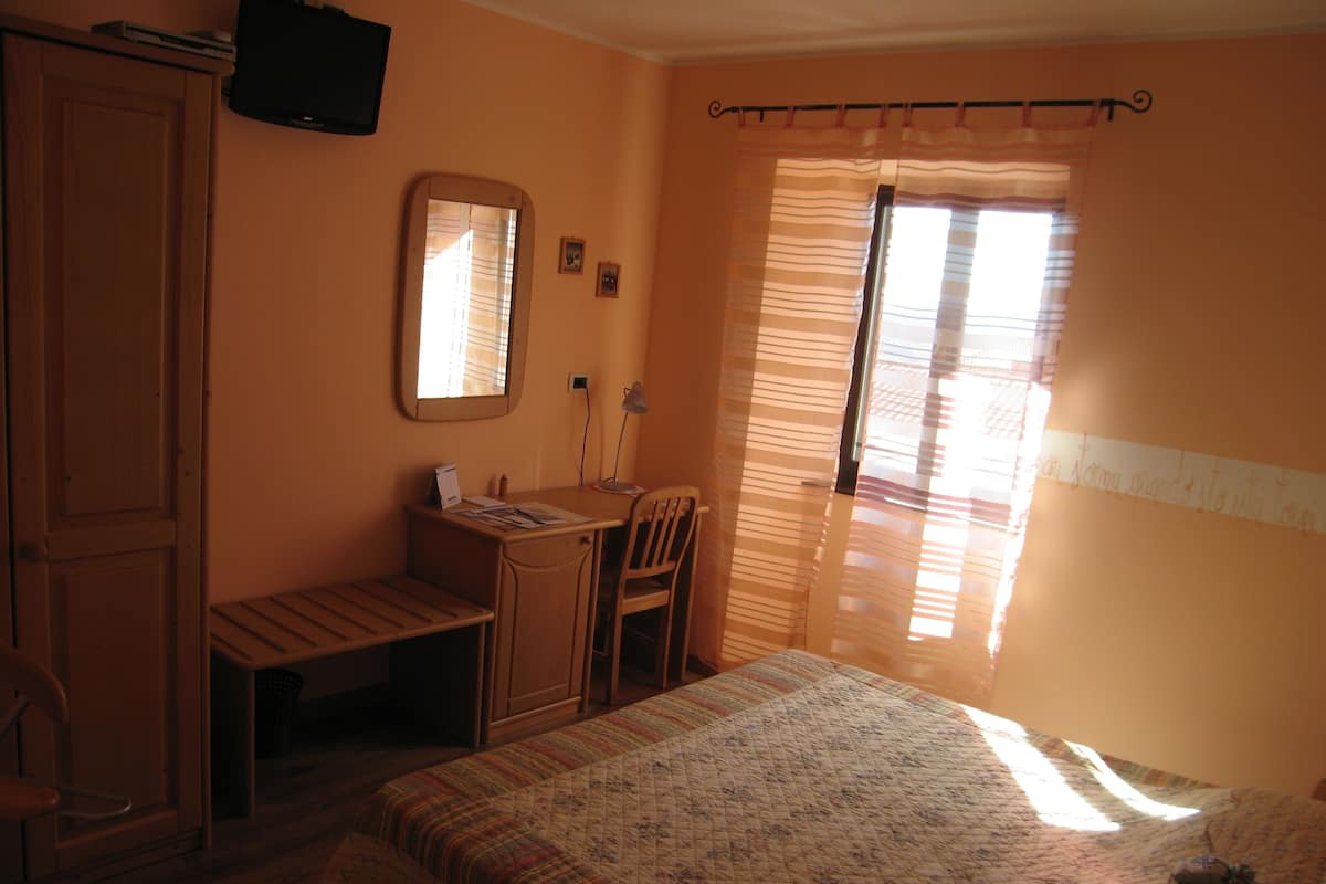Quarto com cama, escrivaninha, cadeira, guarda-roupa, janela com cortinas e televisão na parede.