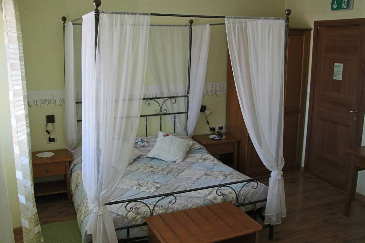 Uma cama de dossel com cortina, uma mesa de cabeceira, um criado-mudo e um banco de madeira.