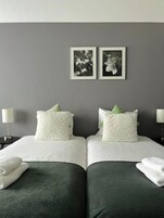 Quarto de hotel com duas camas, uma parede cinzenta e duas imagens emolduradas.