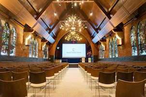 Sala de conferência com uma tela grande na frente, fileiras de cadeiras e janelas com vitrais.