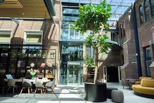Amplo espaço interno com design moderno, apresentando uma área de estar central com mesas e cadeiras, cercada por paredes de tijolo e grandes janelas que permitem a entrada de luz natural.