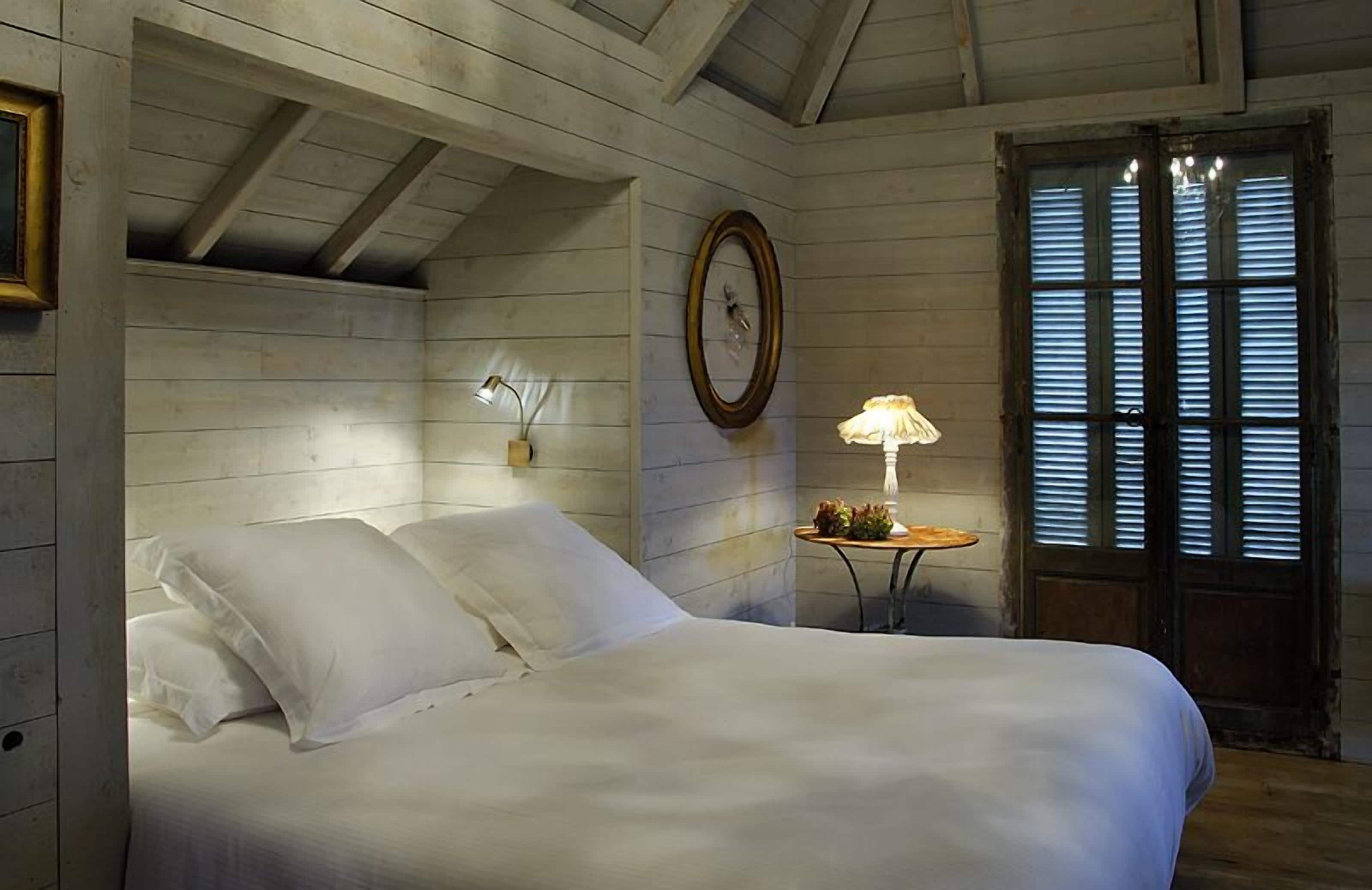 Quarto com teto de madeira, cama com roupa branca, luminária de cabeceira, espelho emoldurado e janela com persianas.