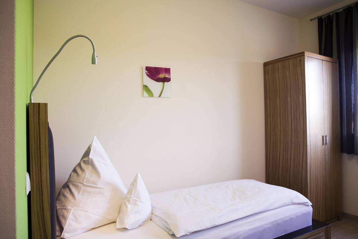 Cama bem arrumada com roupa de cama branca, uma luminária fixada na parede, um guarda-roupa de madeira e uma pequena moldura com uma flor na parede.