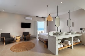 Quarto moderno com cama, duas cadeiras, televisão, banheiro com pia e espelho, e vista para o mar.
