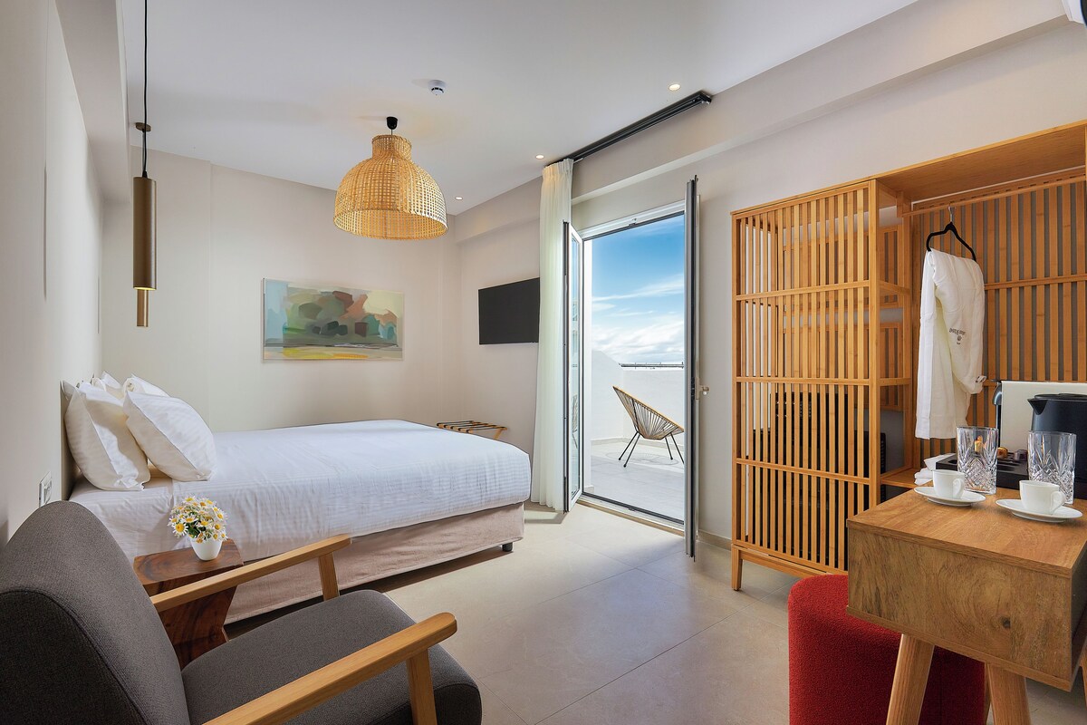 Quarto de hotel moderno com cama, escrivaninha, cadeira, luminária pendente e porta de vidro deslizante que dá para uma varanda.