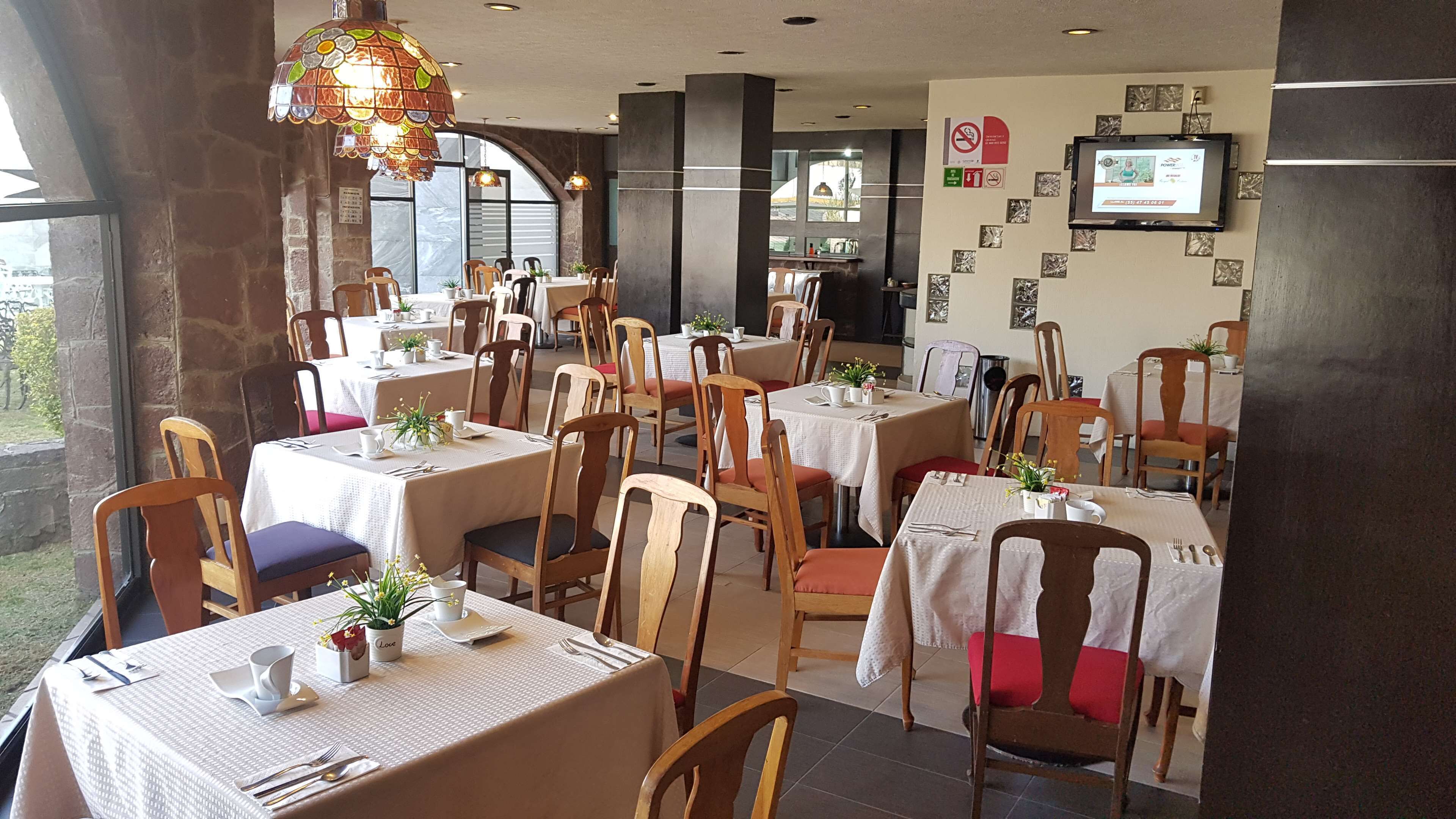 Restaurante com várias mesas e cadeiras, algumas decoradas com plantas. Há placas nas paredes e uma televisão montada.