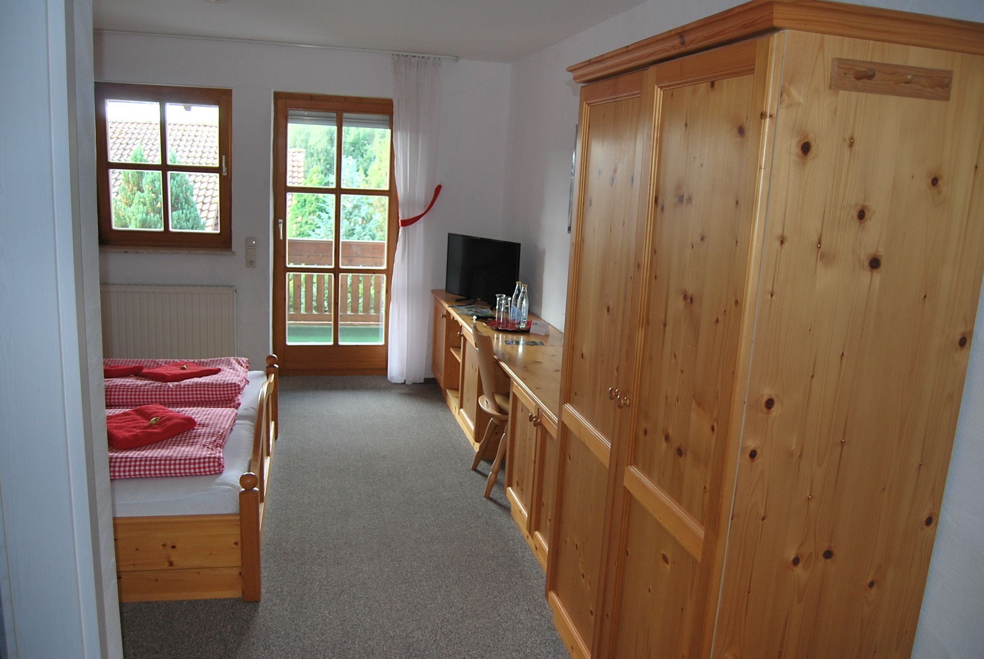 Quarto com cama, guarda-roupa de madeira, escrivaninha com televisão e janela com vista para áreas verdes.