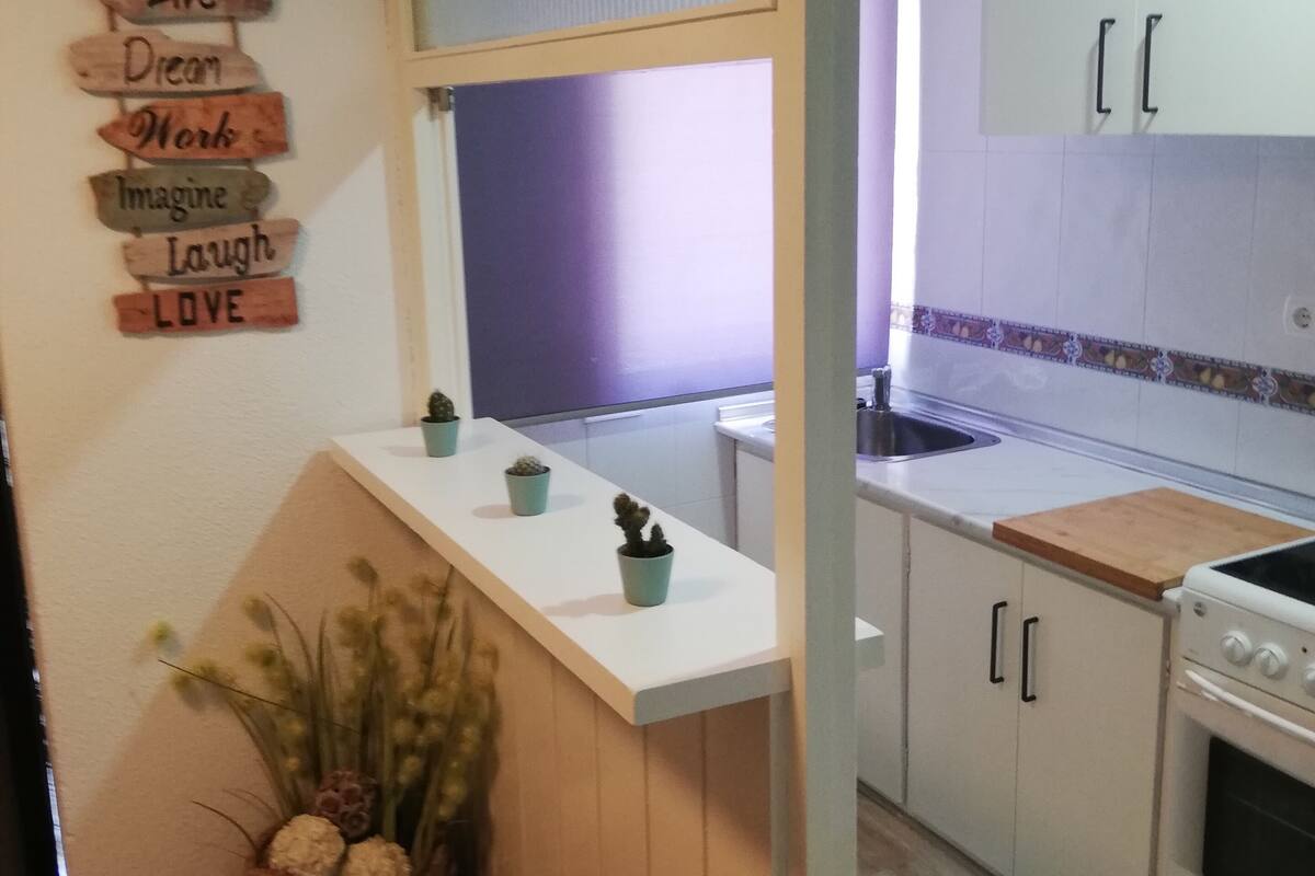 Cozinha com bancada branca, armários de madeira e uma pequena janela. Há uma decoração de parede com frases motivacionais e uma cesta com plantas.