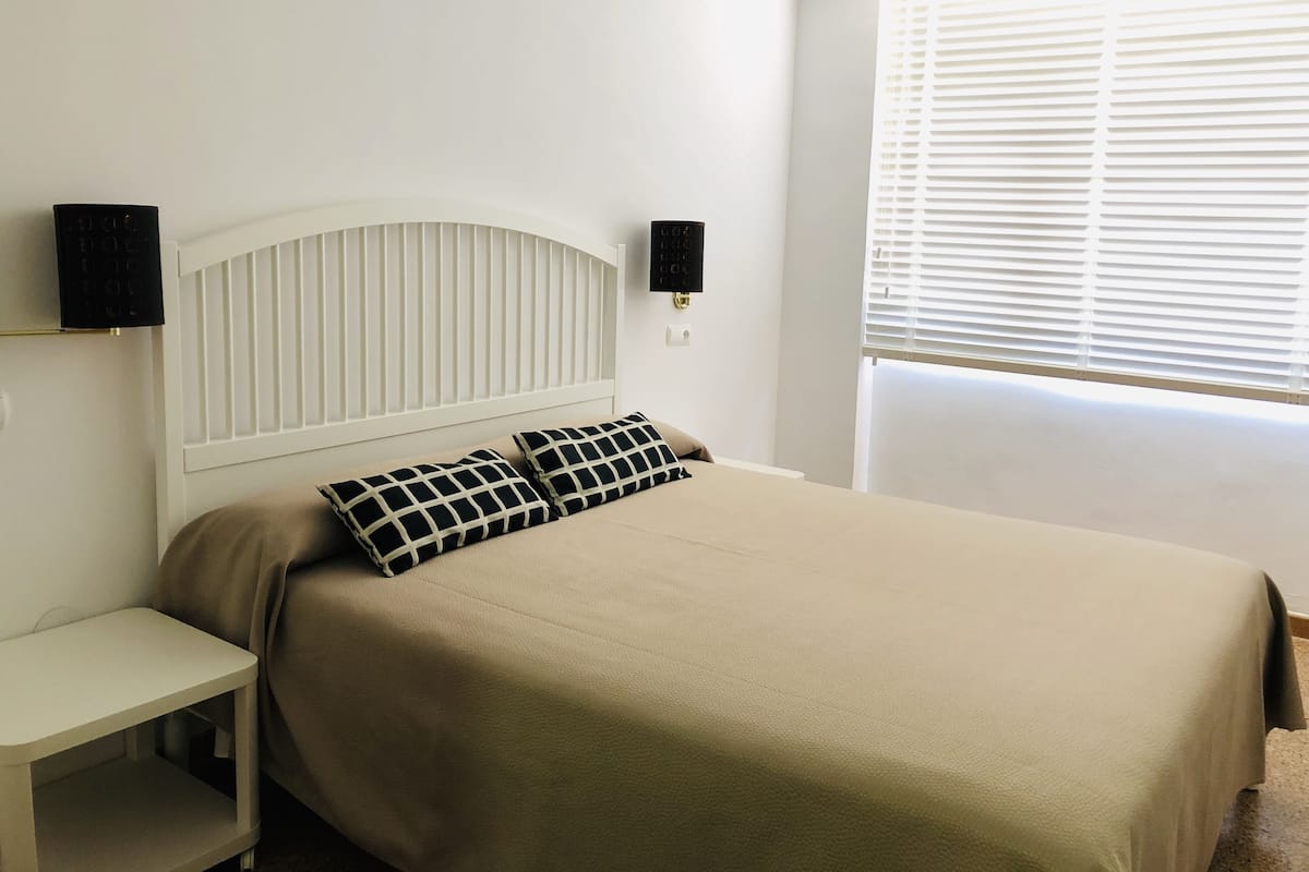 Quarto com cabeceira branca, duas almofadas com padrão preto e branco, colcha bege, mesa de cabeceira branca e janela com persianas.