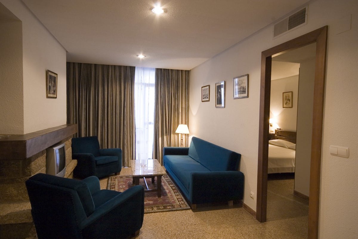 Quarto de hotel com um sofá azul, duas poltronas, uma mesa pequena e uma televisão.