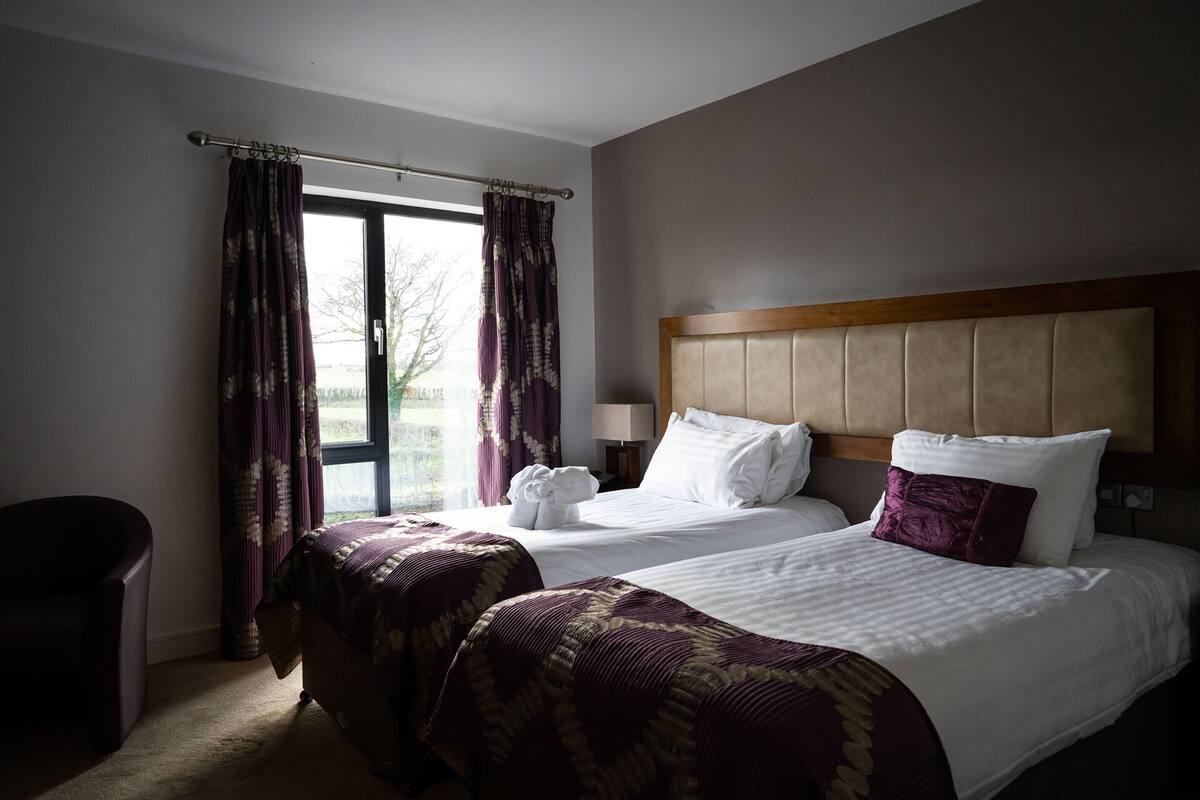 Quarto de hotel com duas camas, janela com cortinas, mesa de cabeceira com abajur e uma cadeira.