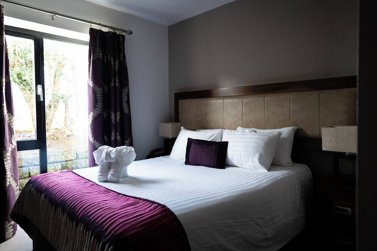 Quarto de hotel com uma cama grande, mesa de cabeceira, abajur e janela com cortinas.