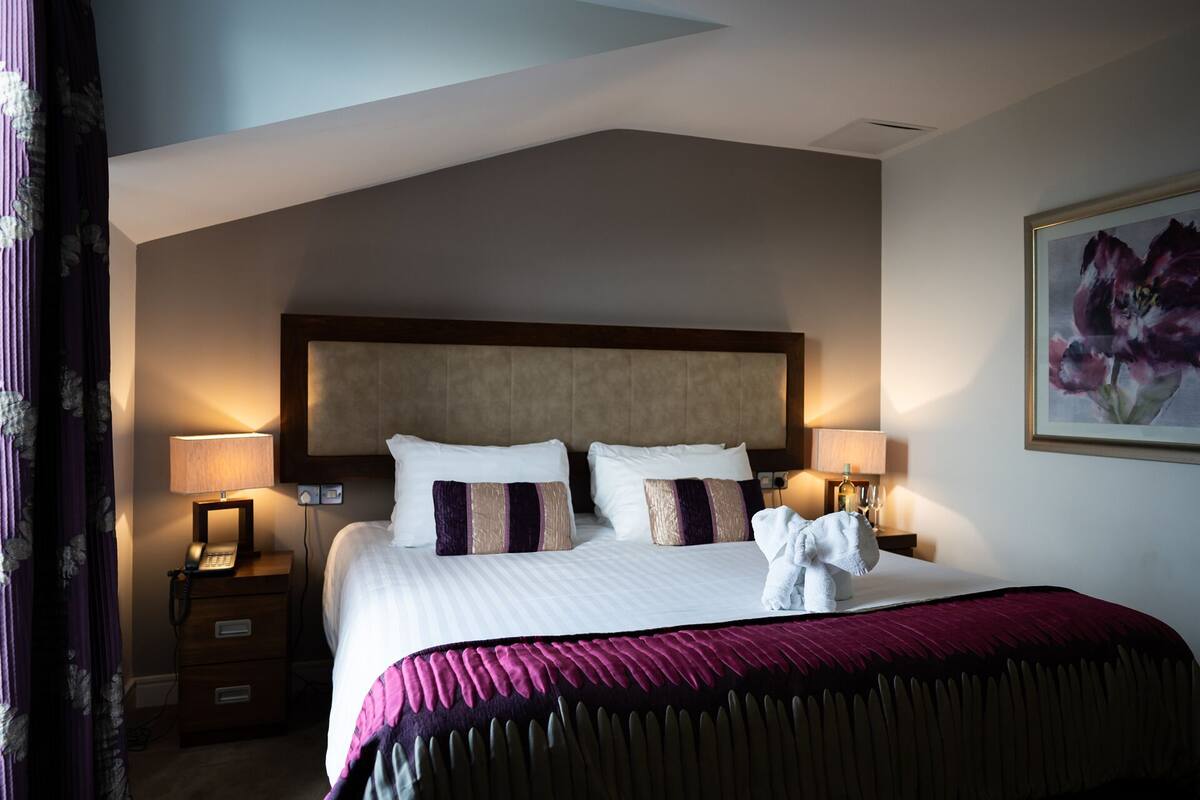 Quarto de hotel com uma cama grande, duas luminárias de cabeceira, um telefone, uma mesa de cabeceira e uma obra de arte floral emoldurada na parede.