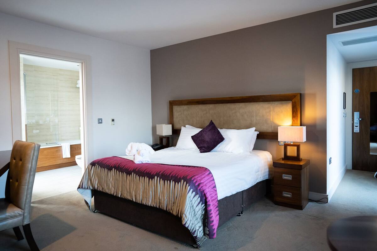 Quarto de hotel com uma cama grande, uma mesa de cabeceira com luminária, uma cadeira e vista para o banheiro.
