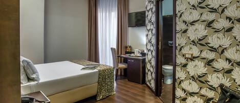 Quarto de hotel com cama, escrivaninha, cadeira e sanita.