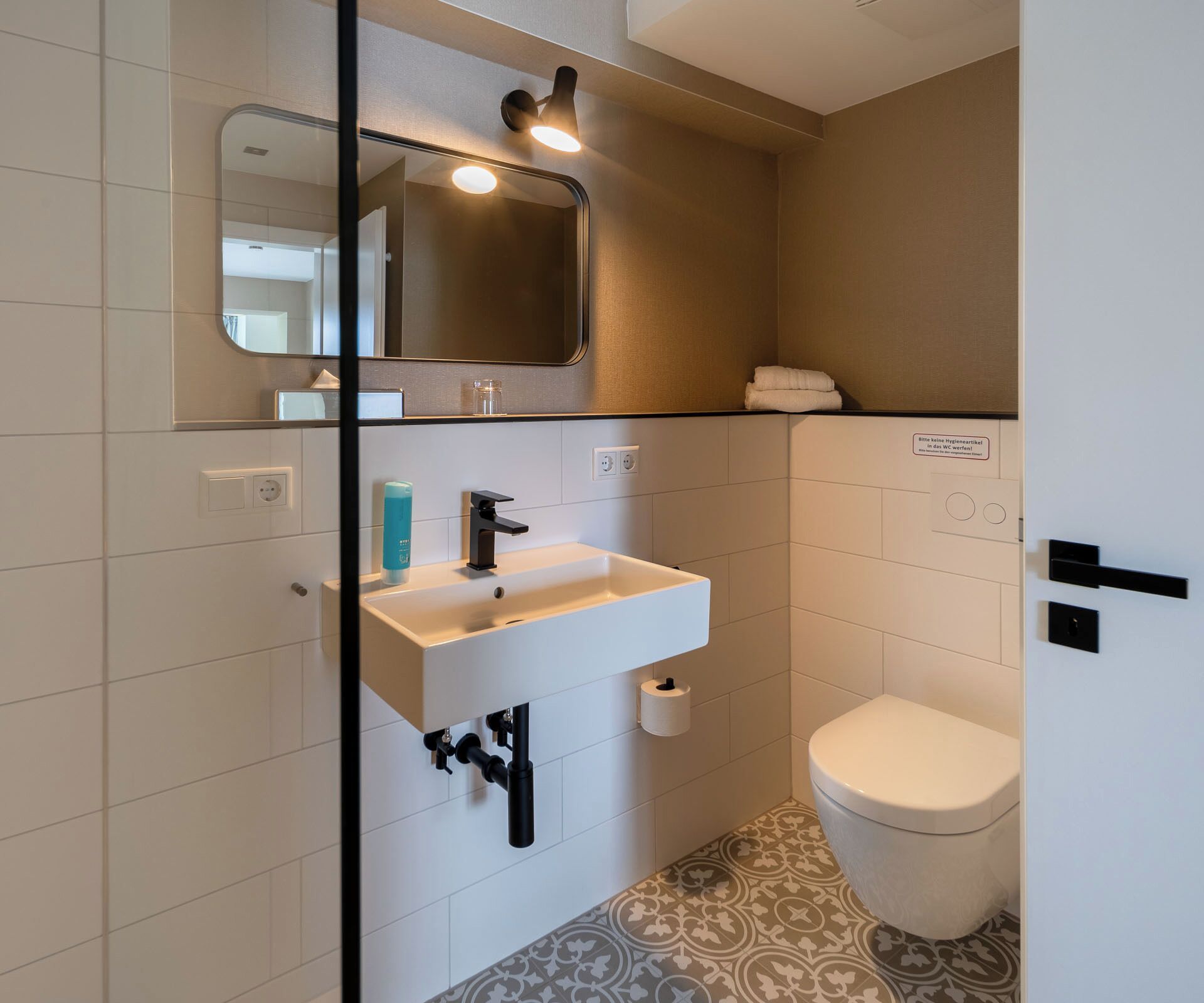 Um banheiro moderno com pia branca, torneira preta e vaso sanitário. Há um espelho sobre a pia e um porta-toalhas acima do vaso.