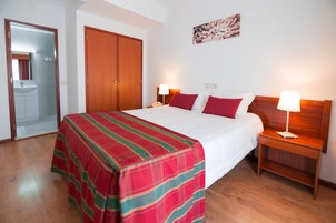 Quarto de hotel com cama, mesas de cabeceira de madeira, colcha de cama listrada em vermelho e verde, guarda-roupa de madeira, banheiro com espelho e pia, e uma obra de arte floral na parede.