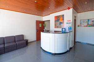 Uma área de recepção com teto de madeira, balcão de recepção, sofá cinza e parede com cartazes informativos.