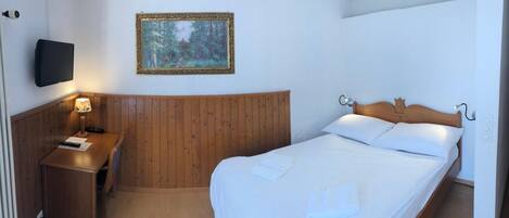 Quarto de hotel com cama de cabeceira em madeira, escrivaninha com luminária, quadro emoldurado na parede e televisão de tela plana na parede.