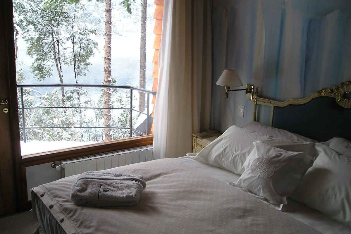 Quarto com cama, janela com vista para árvores, mesa de cabeceira e luminária.
