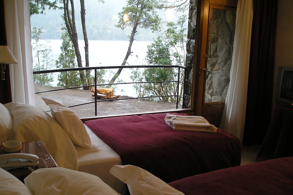 Quarto de hotel com cama, cadeira, mesa e telefone, e vista para um lago através de uma varanda.