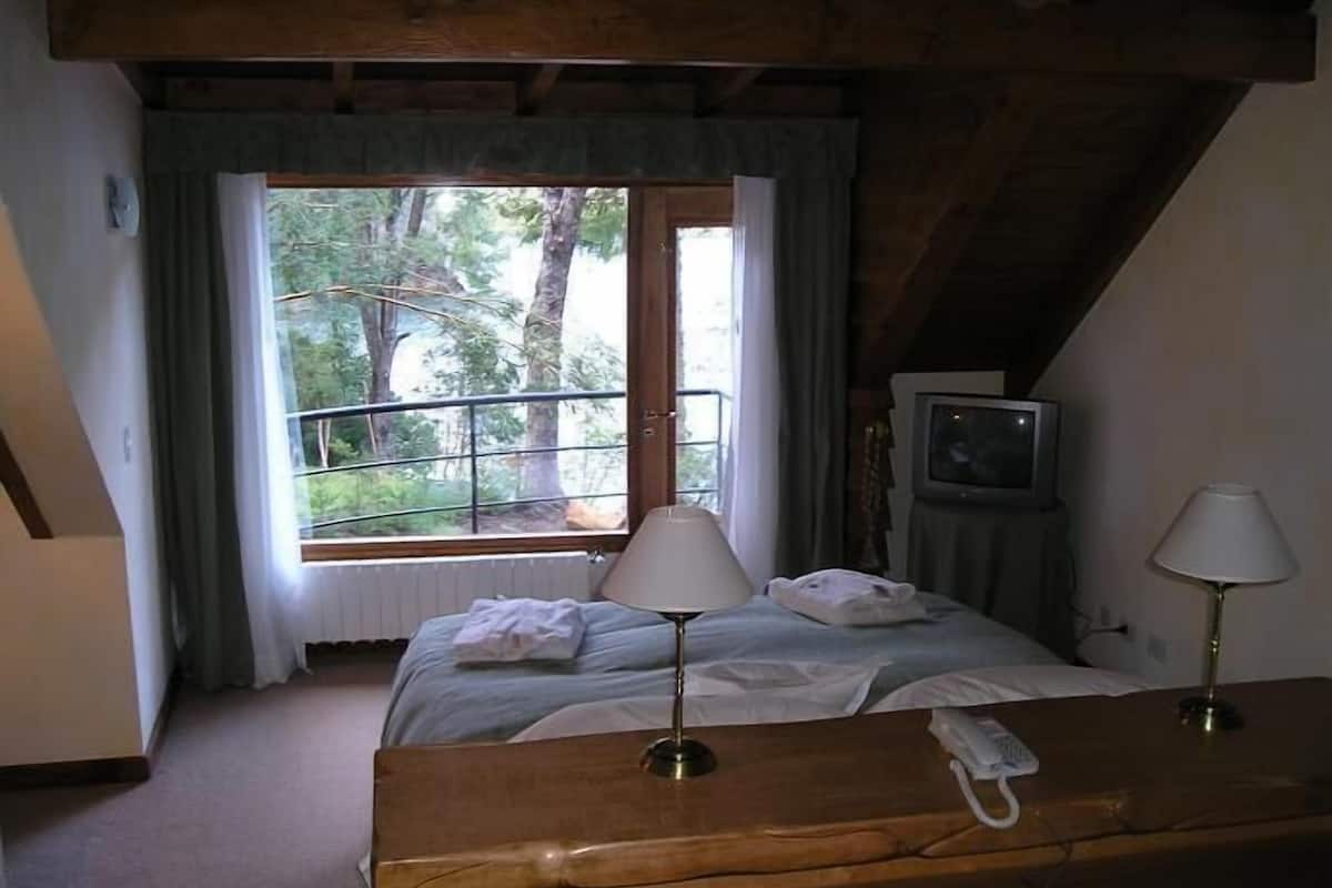 Quarto com cama, escrivaninha de madeira, telefone, televisão e janela com vista para árvores.