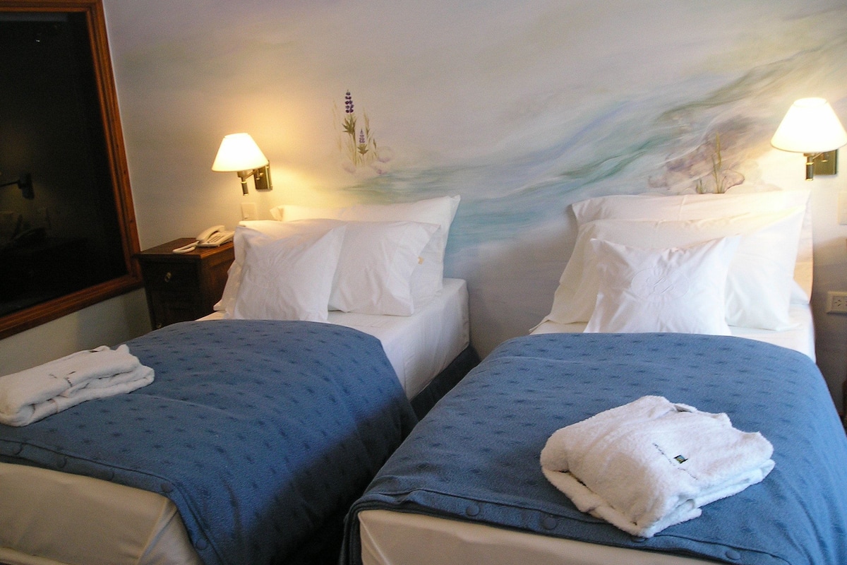Duas camas de casal com roupa de cama branca e colchas azuis, cada uma com uma toalha branca dobrada por cima. Há luminárias fixadas na parede de cada lado das camas, e um quadro com uma paisagem marítima na parede atrás das camas.