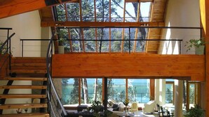 Um amplo espaço estilo loft com escada de madeira, grandes janelas com vista para o oceano e uma área de estar acolhedora com sofás e uma mesa de centro.