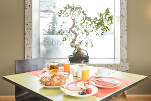 Mesa de café da manhã com croissants, ovos e suco de laranja, com vista para uma janela e uma árvore bonsai.