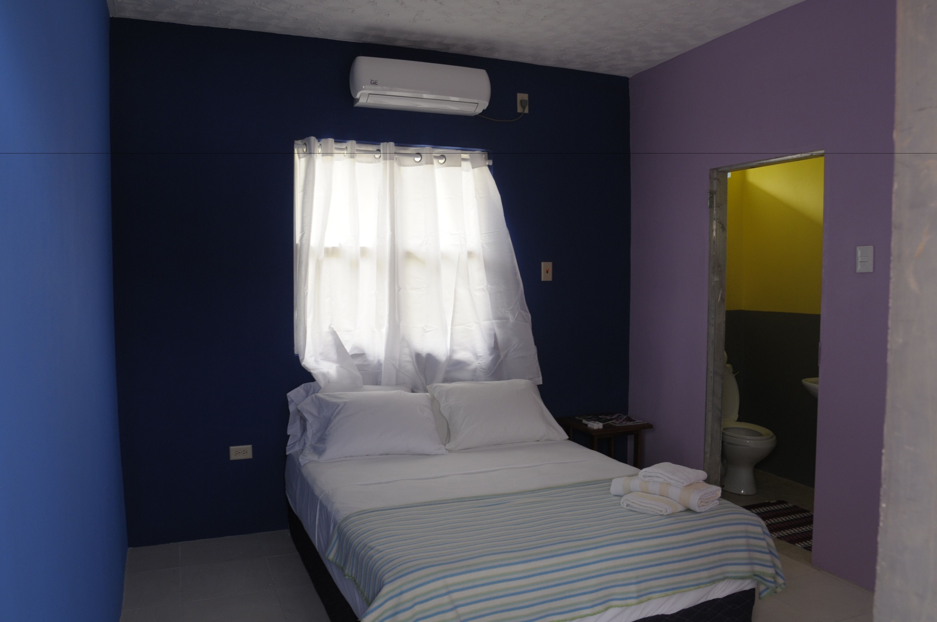 Quarto com cama, cortinas brancas, mesa de cabeceira e banheiro visível através de uma porta aberta.