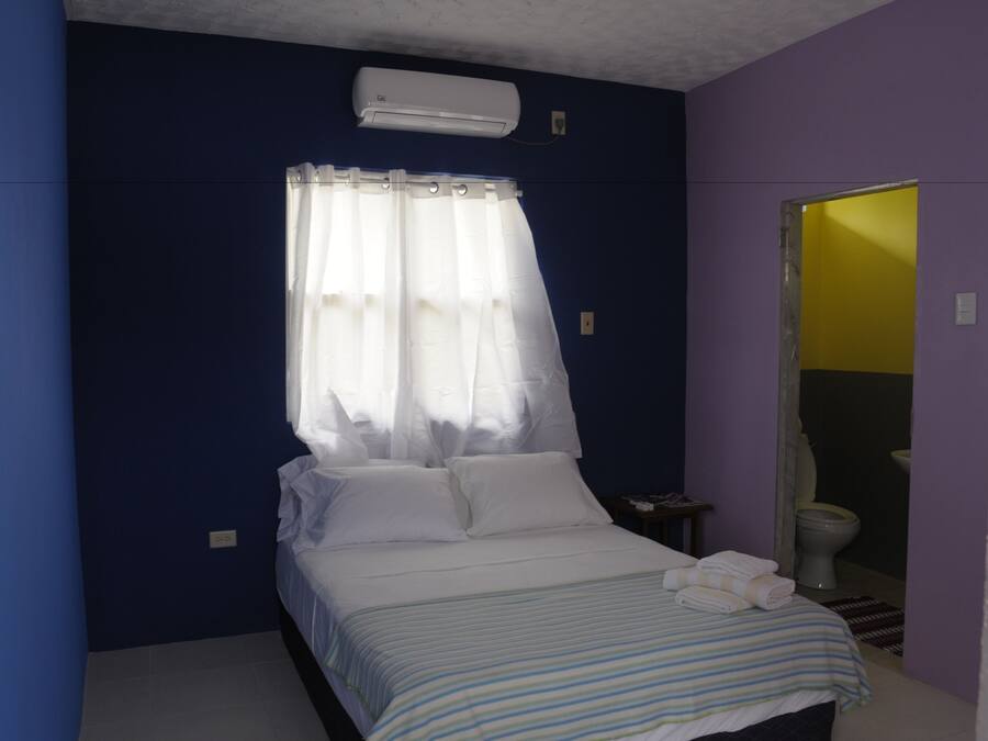 Quarto com cama, cortinas brancas, mesa de cabeceira e banheiro visível através de uma porta aberta.
