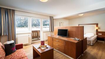 Quarto de hotel com escrivaninha de madeira, televisão, sofá, cama e vista para as montanhas pelas janelas.