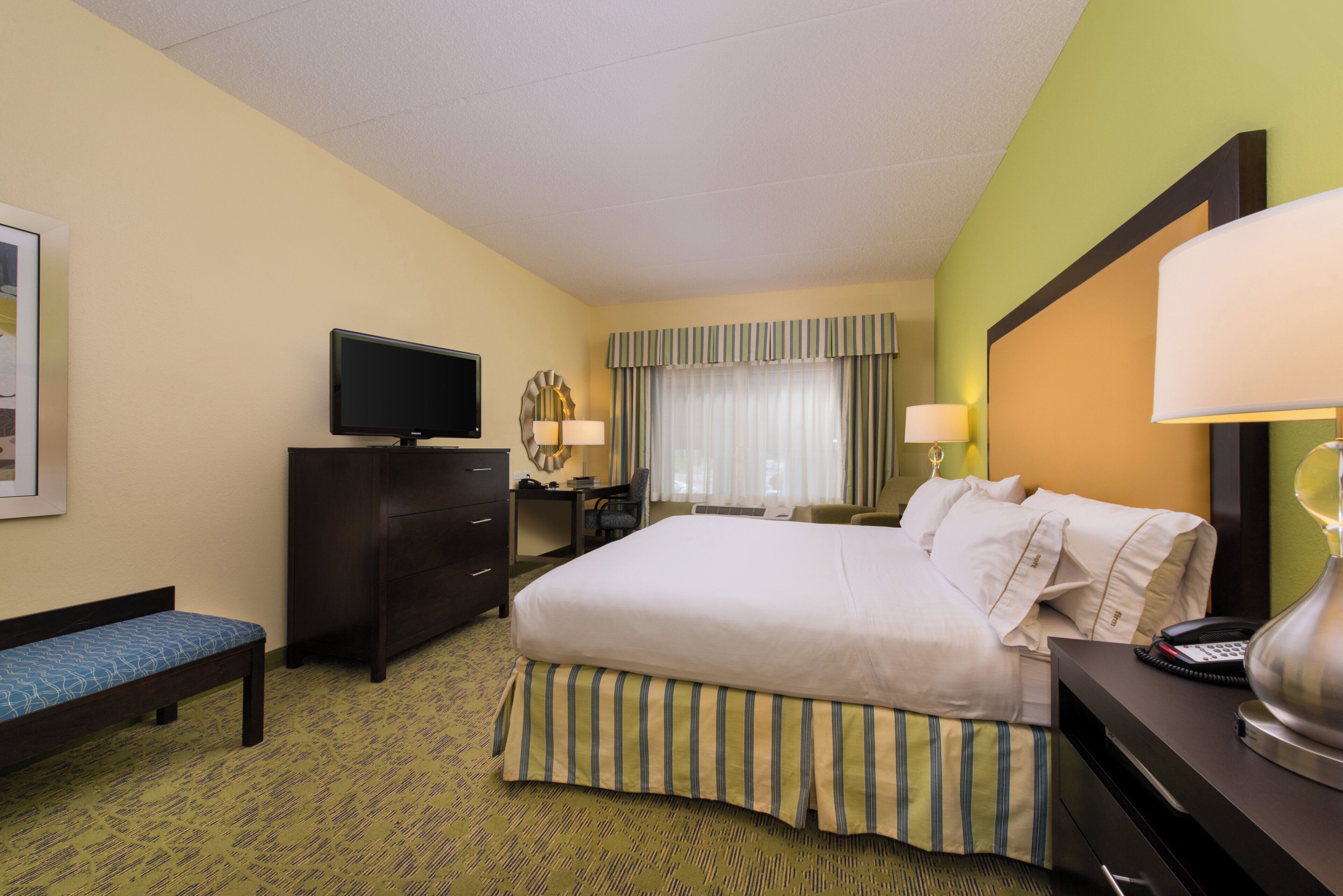 Quarto de hotel com cama, escrivaninha, televisão, abajur e cadeira.