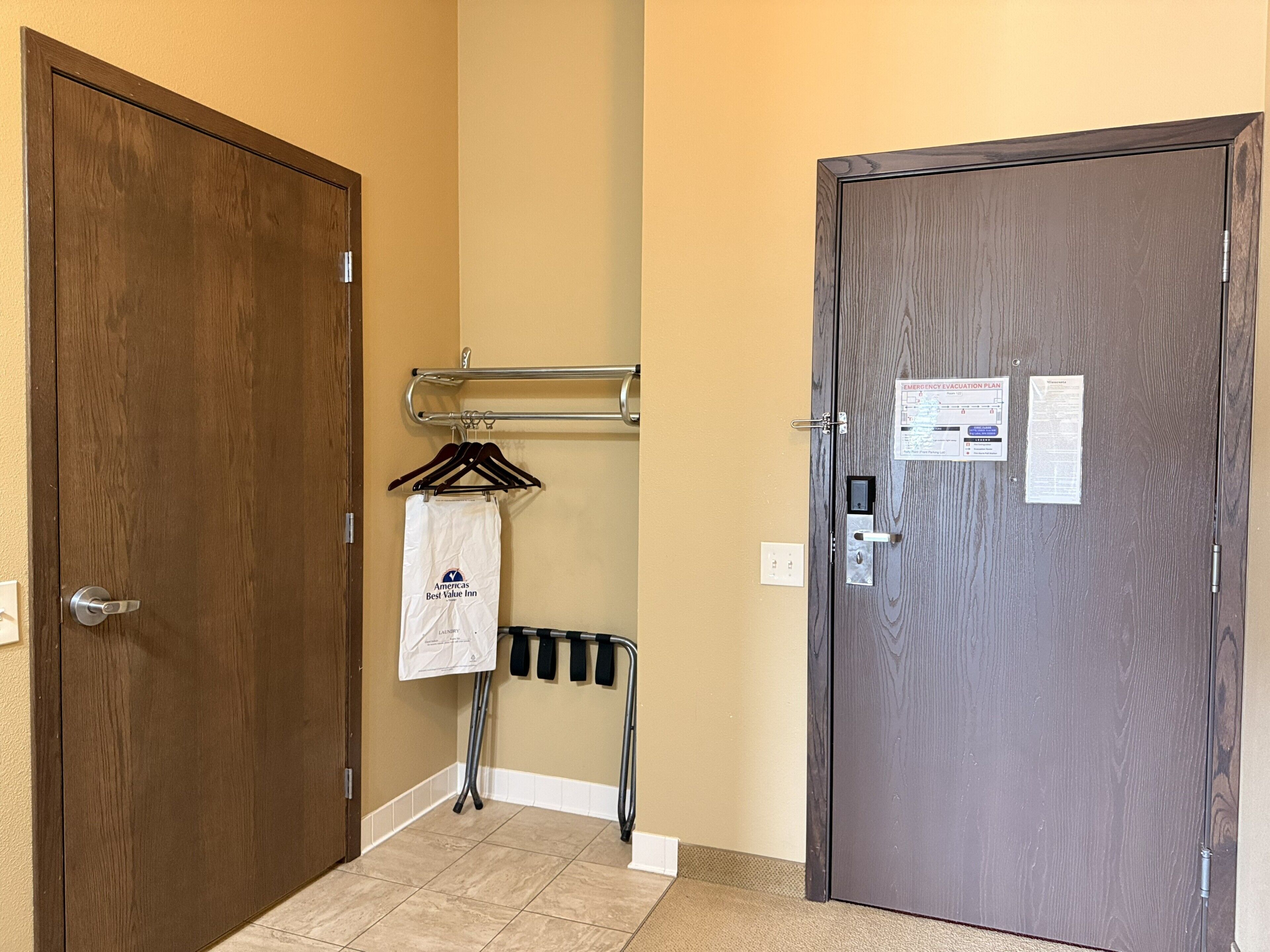 Quarto de hotel com duas portas de madeira, um porta-toalhas com uma toalha branca, outro porta-toalhas de metal e um pequeno banco preto.